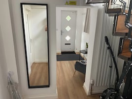 Um corredor com espelho, porta com janelas em formato de losango, mesa pequena com planta e aspirador de pó.
