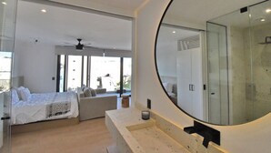 Quarto moderno com uma cama grande, um sofá, box de vidro para o chuveiro e um espelho amplo.