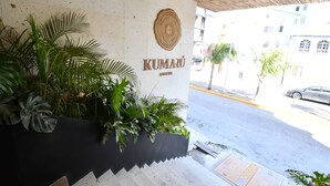 Entrada de hotel com uma placa indicando "KUMARÚ SUITES" e um emblema decorativo. Há plantas em vasos e uma escadaria que leva à entrada.