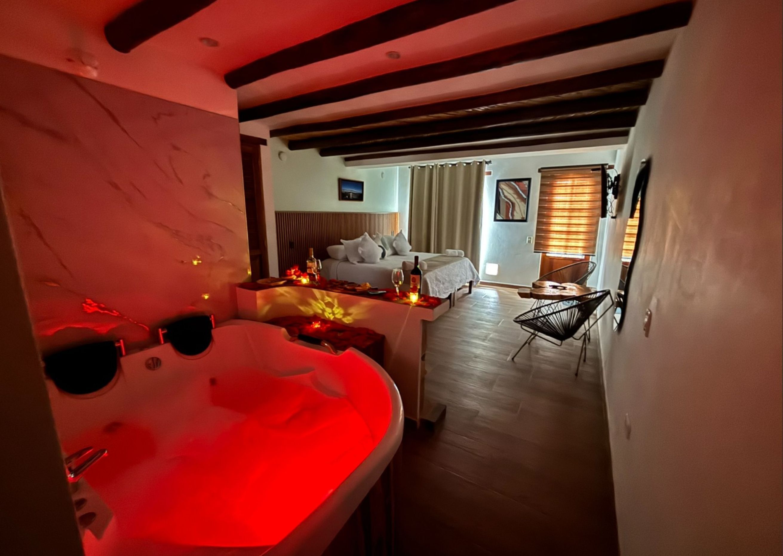 Quarto com cama, uma mesinha com velas, uma cadeira e vista para outro quarto com cama e cortinas.