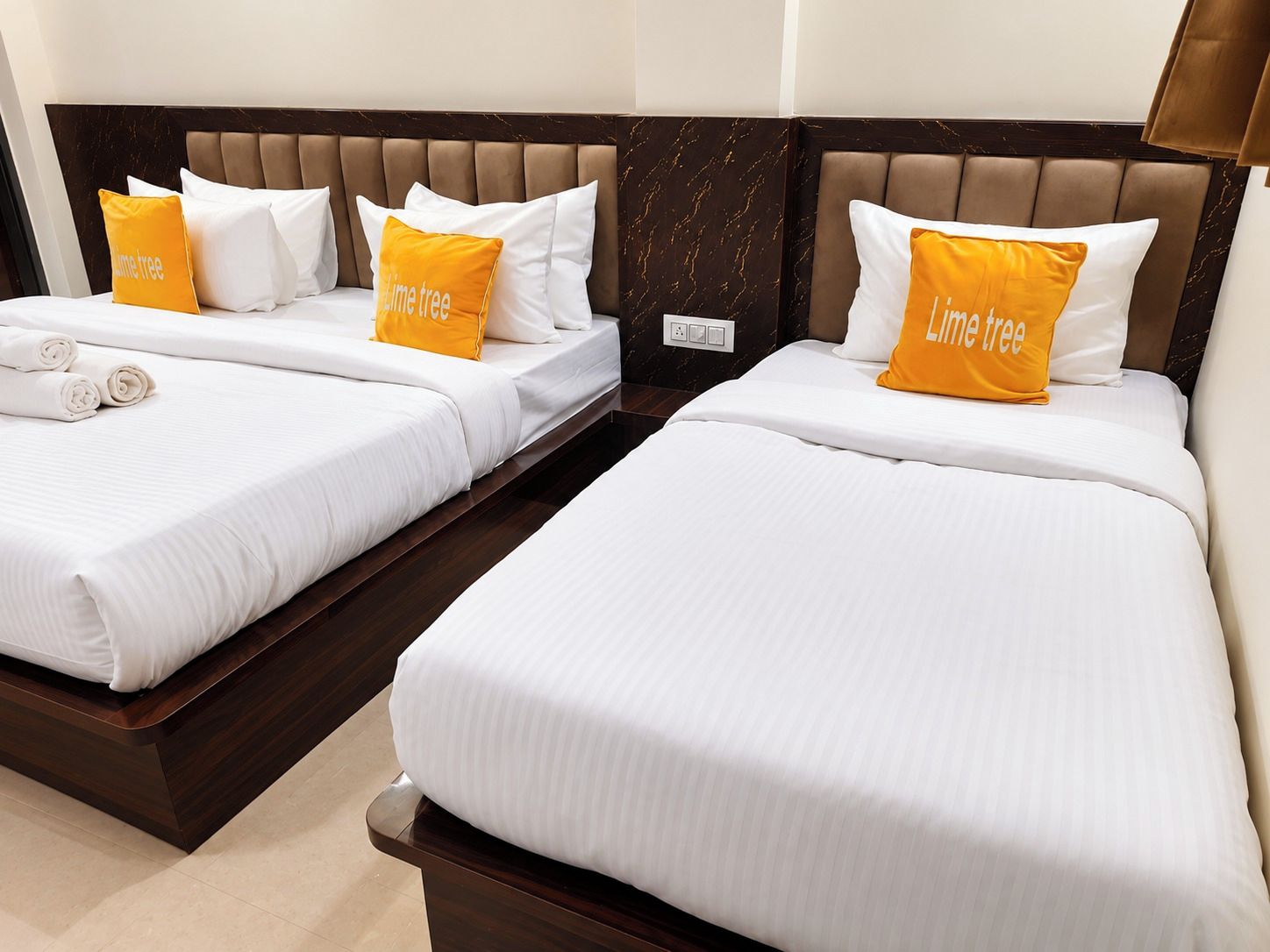 Duas camas de casal com roupa de cama branca e almofadas laranja, uma cabeceira escura e uma parede com padrão marmorizado.