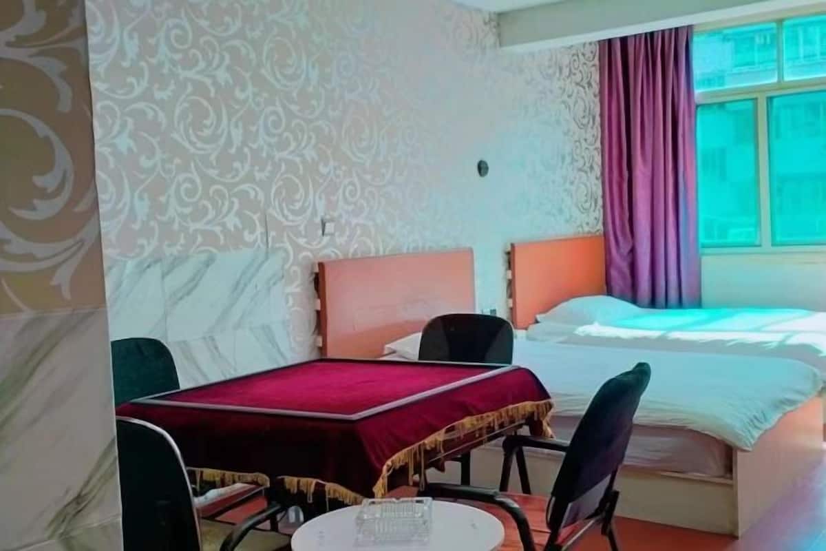 Quarto de hotel com uma cama, duas cadeiras, uma mesinha e uma janela com cortinas.