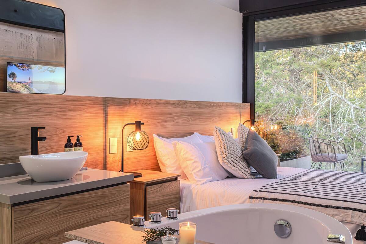 Quarto moderno com uma cama grande, mesa de cabeceira e vista para árvores pela janela.