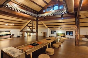 Um salão espaçoso com teto alto e vigas de madeira, com uma mesa comprida e banquetas, e uma tela de televisão.