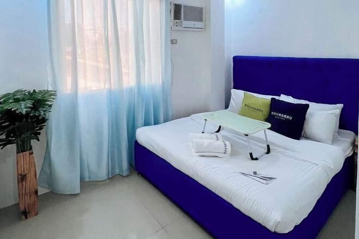 Um quarto com cama, mesa de cabeceira, planta em vaso e janela com cortinas.