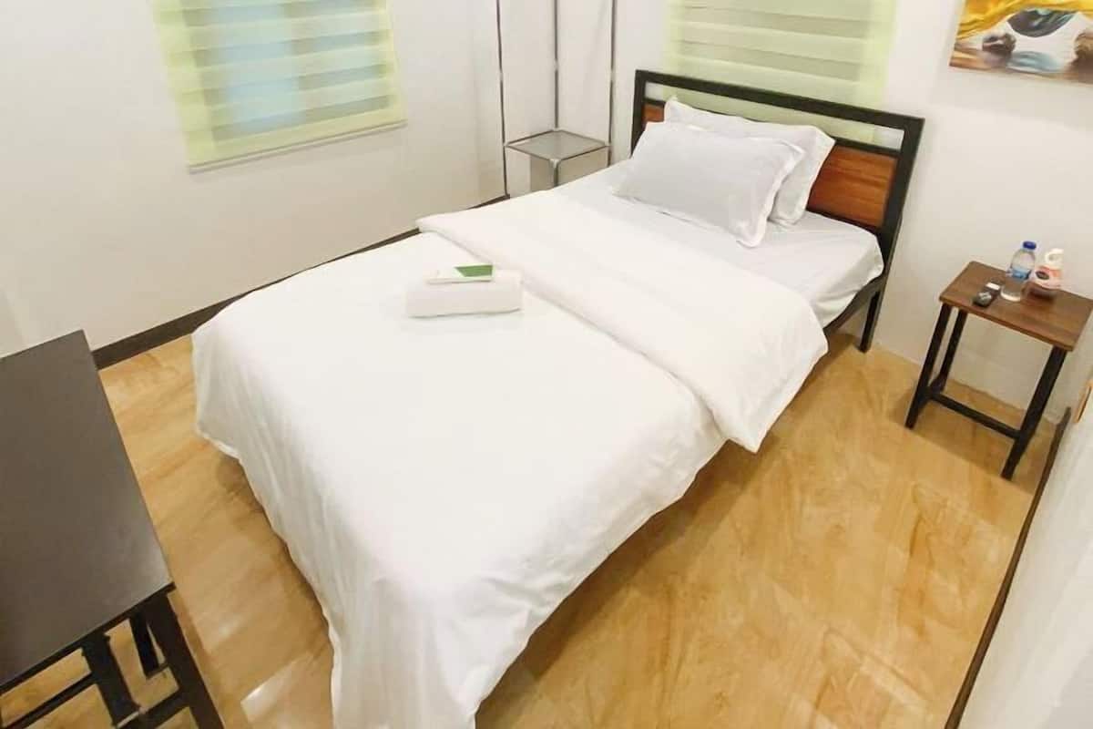Uma cama bem arrumada com roupa de cama branca, uma pequena mesa de cabeceira com uma garrafa e uma caixa de lenços, e uma cadeira em um quarto com piso de madeira e uma janela com persianas.