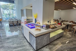 Espaço de refeições moderno com piso de mármore, balcão de buffet com diversos alimentos e vista para o exterior.
