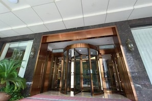 Uma porta giratória com painéis de vidro e estrutura metálica, dando acesso a um edifício com piso de mármore e uma planta em vaso.