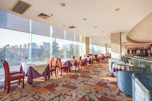 Um espaço de refeições de hotel com mesas, cadeiras, janelas amplas e vista para edifícios.