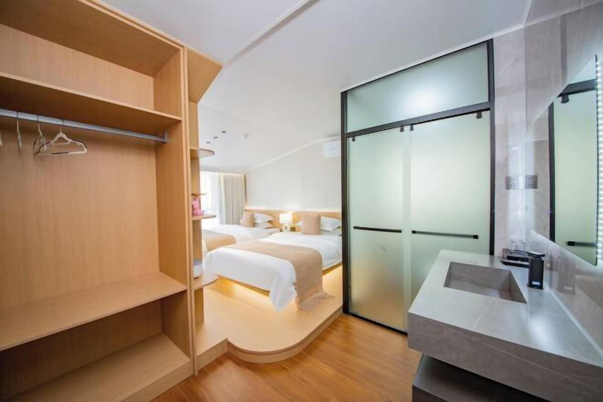 Quarto de hotel moderno com guarda-roupa de madeira, cama com roupa de cama branca, divisória de vidro e banheiro contemporâneo com pia e espelho.