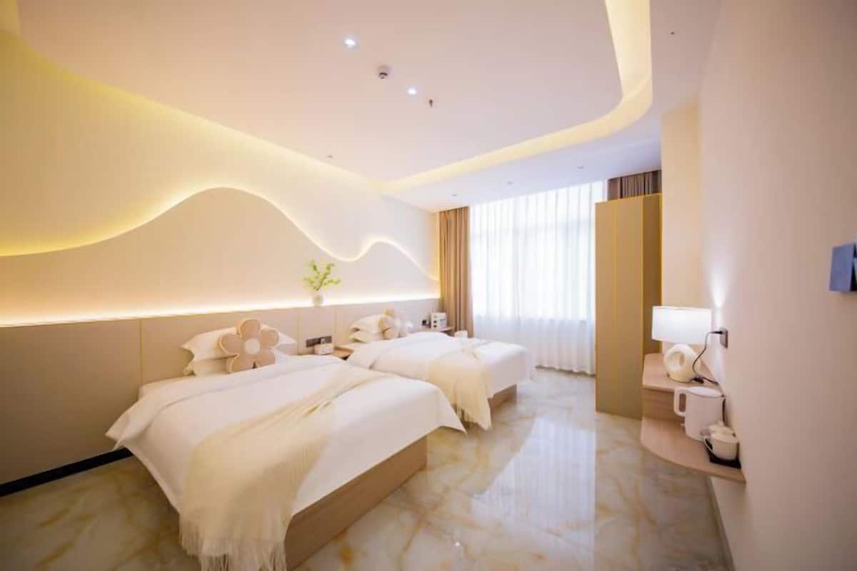 Quarto de hotel moderno com duas camas, uma toucador com espelho e uma janela grande com cortinas.