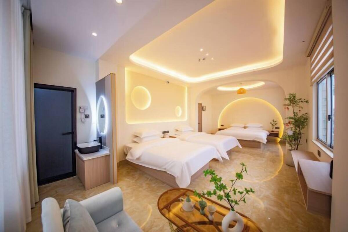 Quarto de hotel moderno com sofá, duas camas, mesa de cabeceira, espelho, banheiro pequeno e uma janela grande.