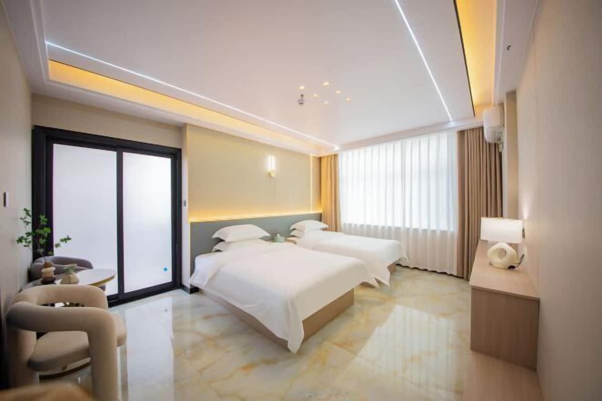 Quarto de hotel moderno com duas camas, uma escrivaninha, uma cadeira e uma janela grande com cortinas.