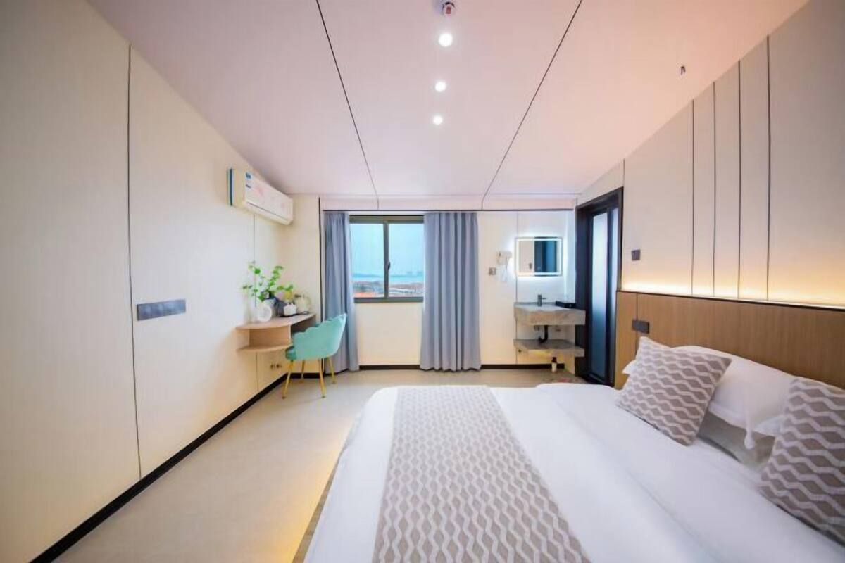 Quarto de hotel moderno com uma cama grande, uma escrivaninha com cadeira, televisão e vista para a cidade pela janela.