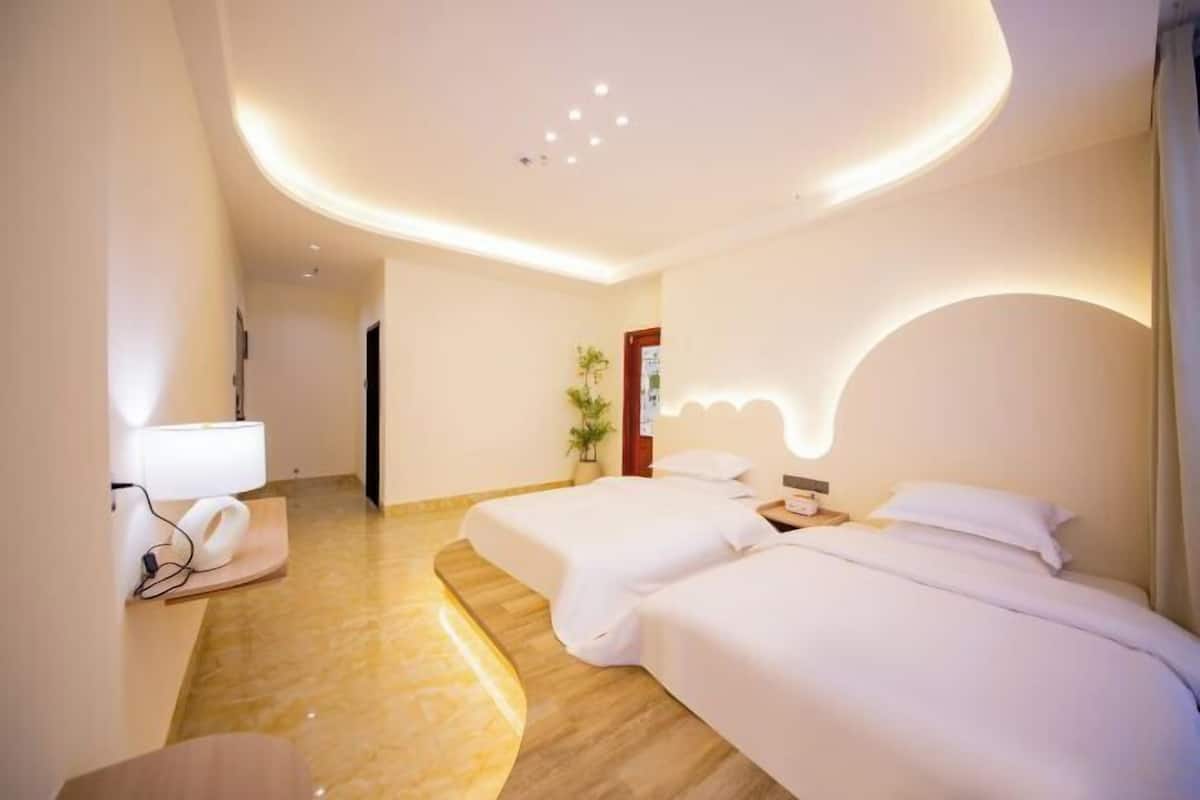 Quarto de hotel com duas camas, uma escrivaninha, uma luminária e uma planta.