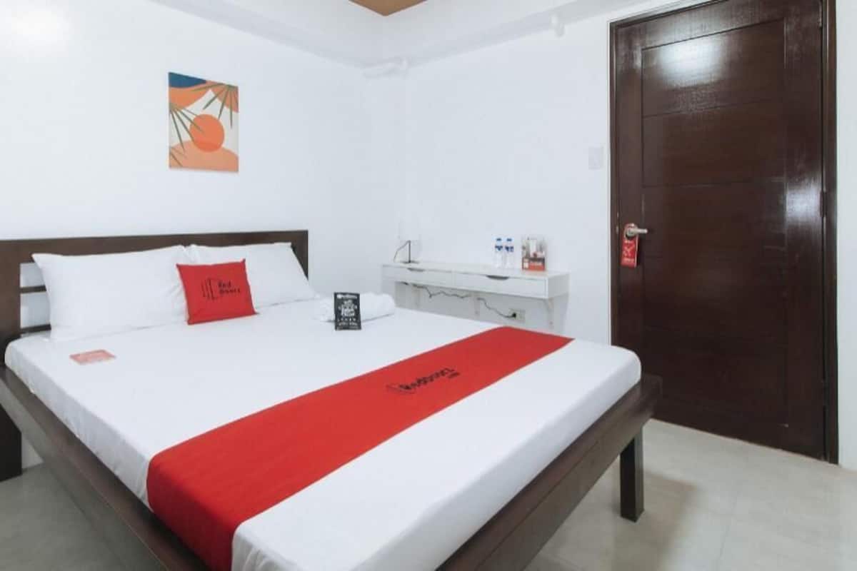 Quarto de hotel com cama, mesa de cabeceira, manta vermelha, pintura na parede e porta de madeira.