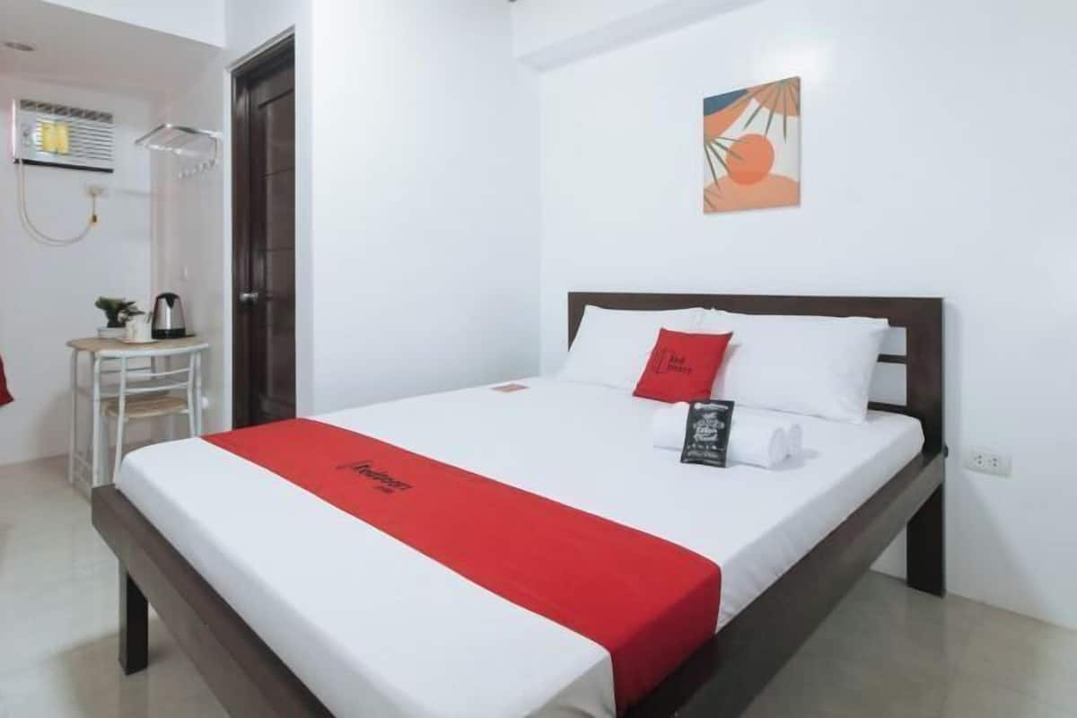 Quarto de hotel com uma cama, uma mesinha com uma cadeira, ar condicionado fixado na parede e um quadro.