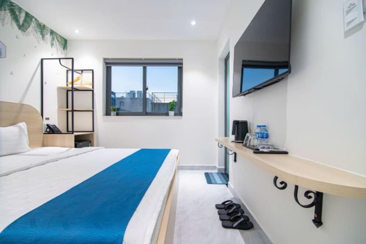 Quarto de hotel com cama, televisão fixada na parede, prateleira pequena com artigos de higiene e janela com vista para edifícios.