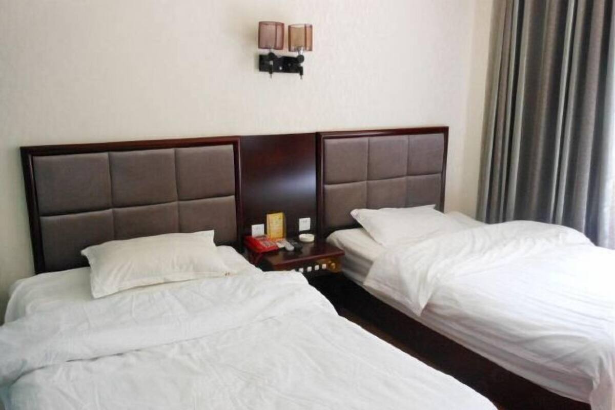 Quarto de hotel com duas camas, uma mesinha de cabeceira e uma luminária na parede.