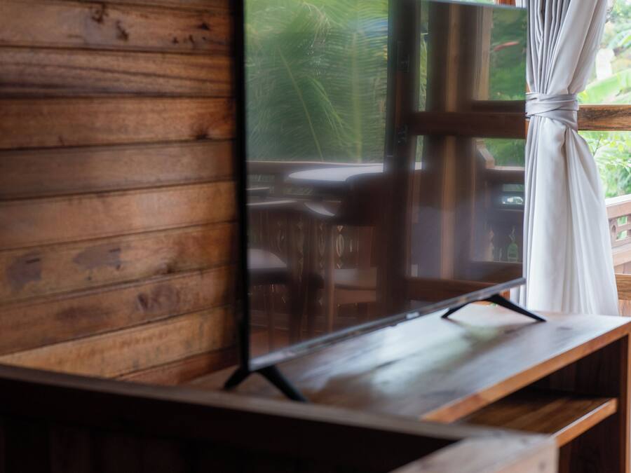 Interior em madeira com televisão em móvel, janela com cortinas e vista para móveis de exterior.