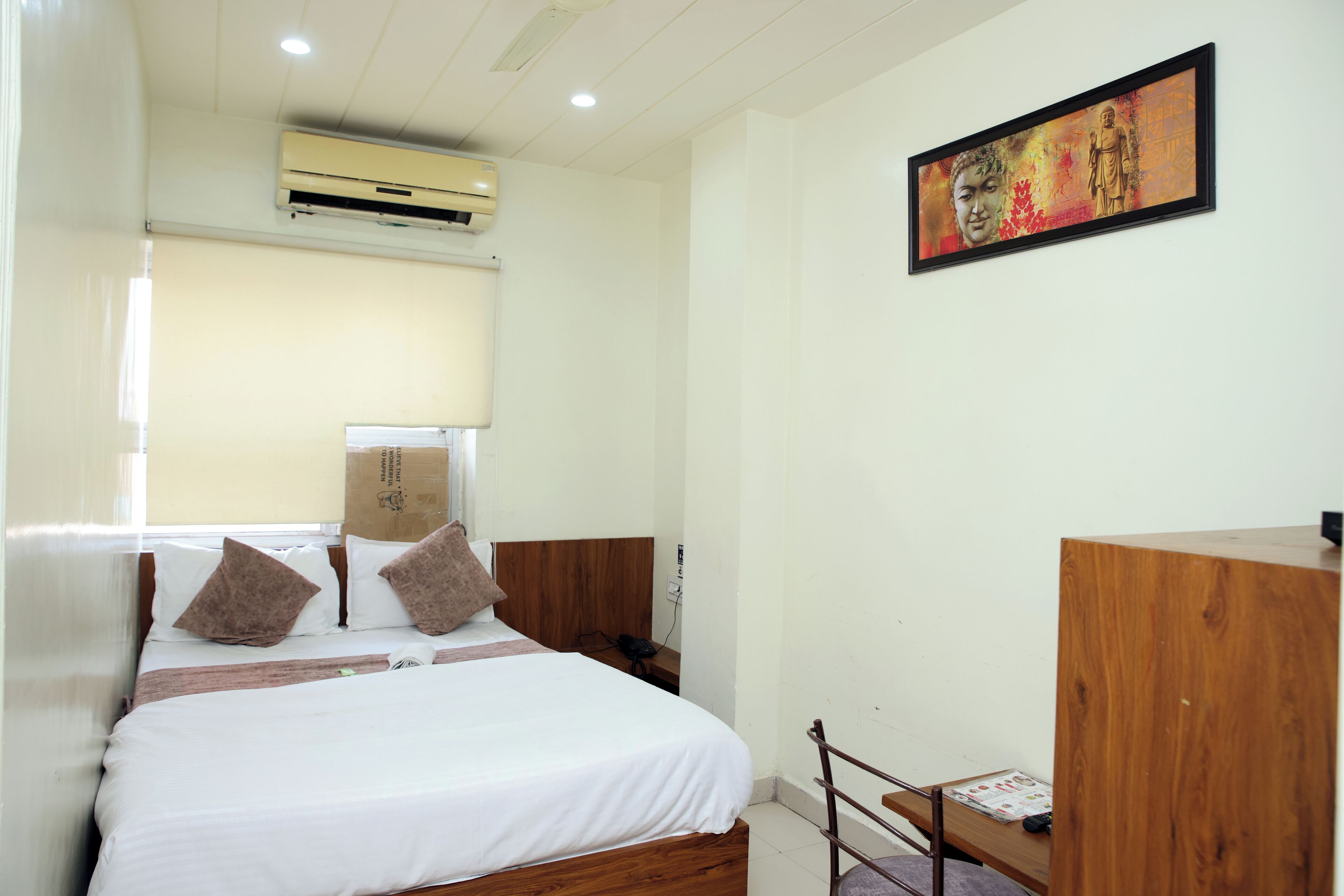 Quarto de hotel com cama, cabeceira de madeira, ar condicionado fixado na parede e uma obra de arte emoldurada acima da cama.