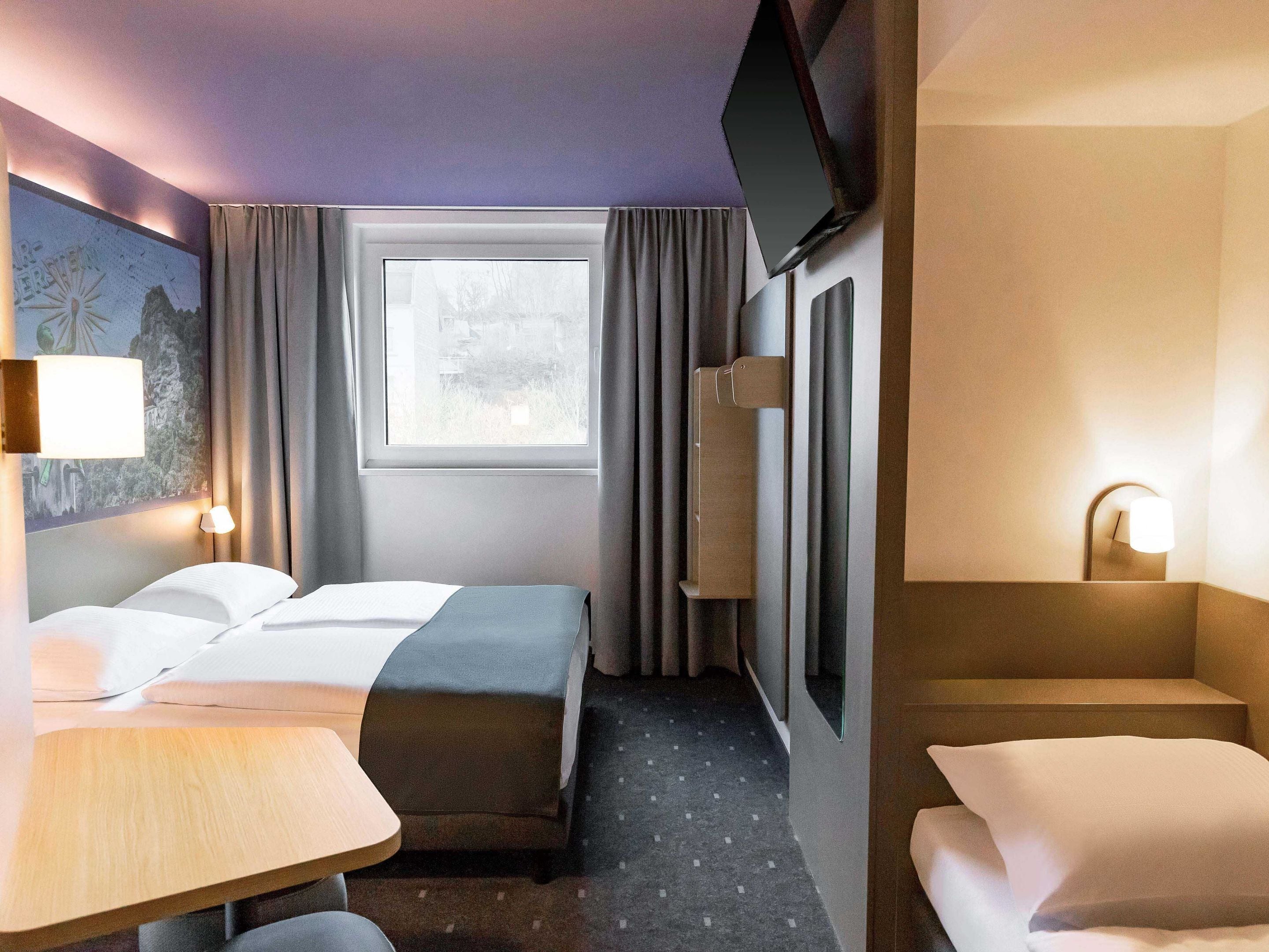 Quarto de hotel com duas camas, uma escrivaninha, uma TV e uma janela com cortinas.