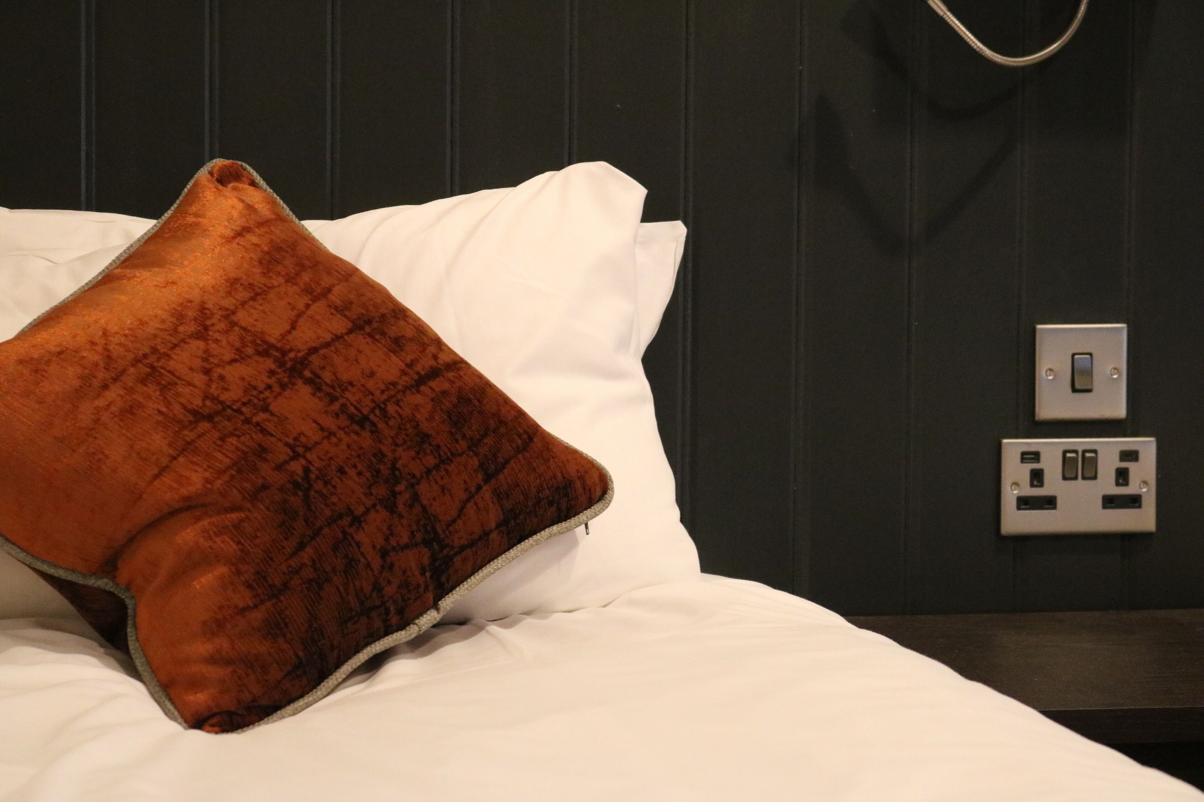 Uma cama com lençóis brancos e travesseiro marrom, uma tomada e um interruptor na parede.