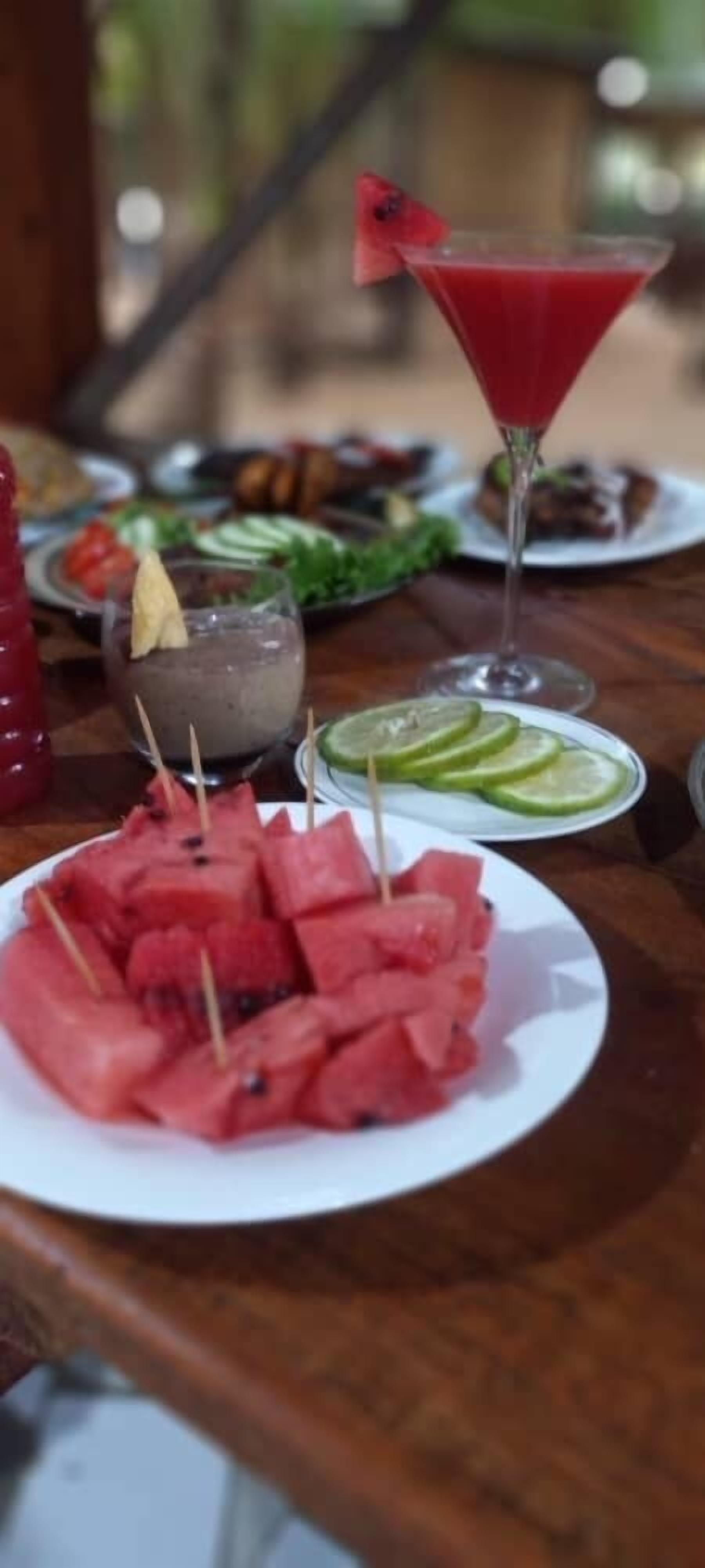 Uma mesa de jantar com espetinhos de melancia, uma taça de martini com uma bebida vermelha e um prato de pepino fatiado.
