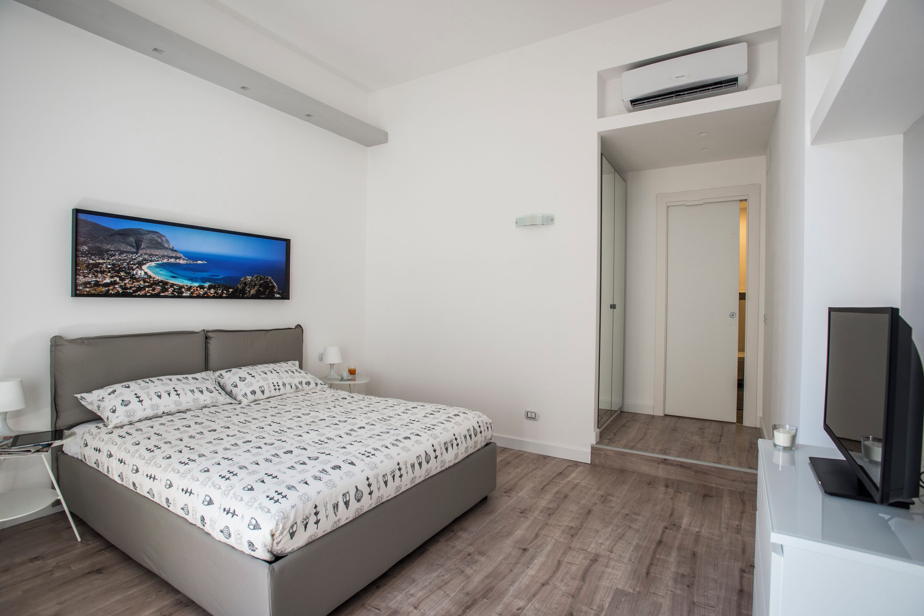 Quarto moderno com cama, televisão e pintura de uma paisagem costeira.