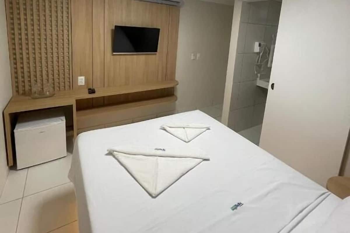 Quarto de hotel com cama, televisão presa a um painel de madeira, guarda-roupa e pequena escrivaninha com luminária.