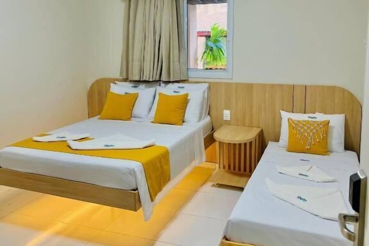 Quarto de hotel com duas camas, uma janela com cortinas, uma pequena mesa de cabeceira de madeira e vista para o verde lá fora.