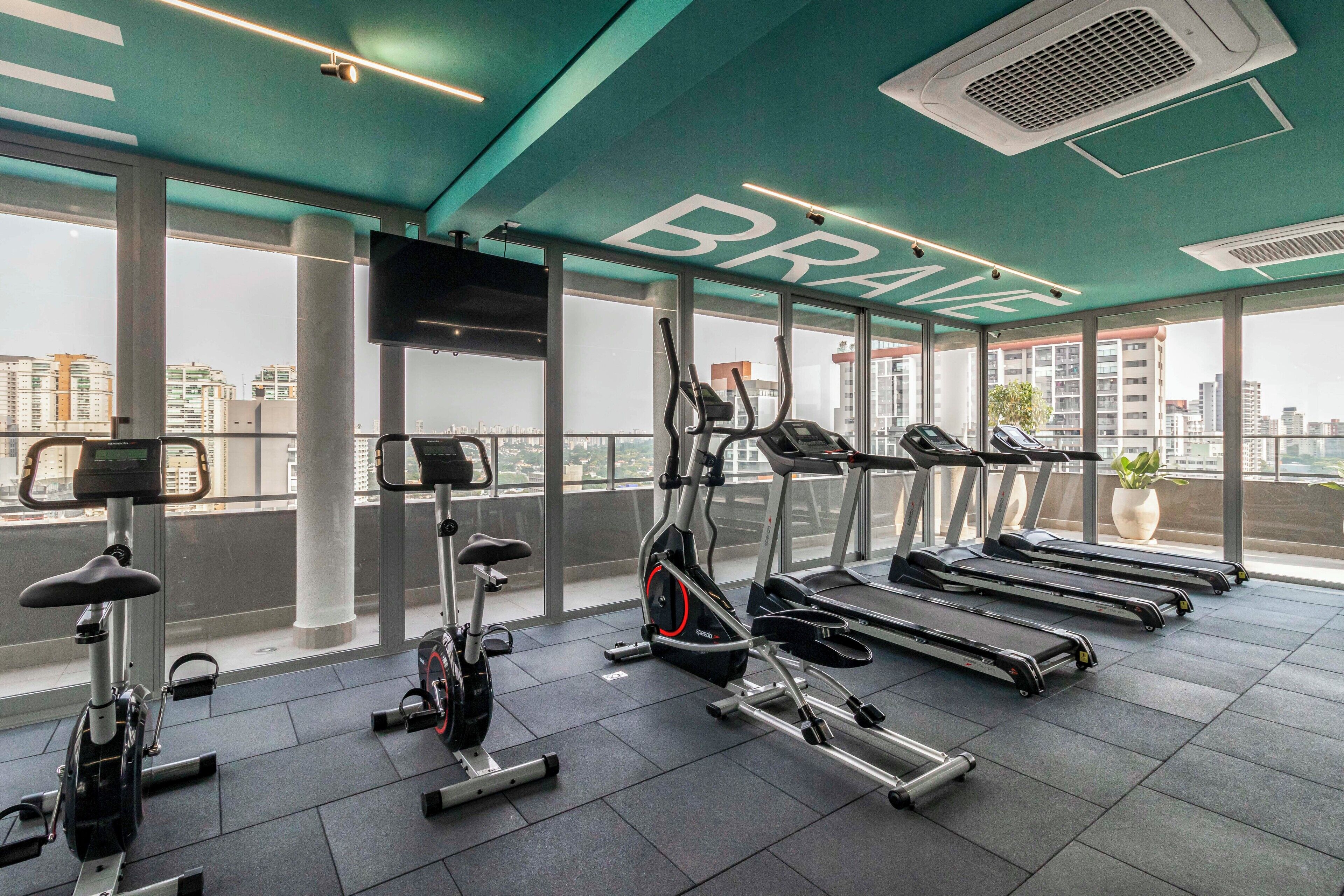 Uma academia com diversos equipamentos de exercício, janelas amplas e vista para a cidade.