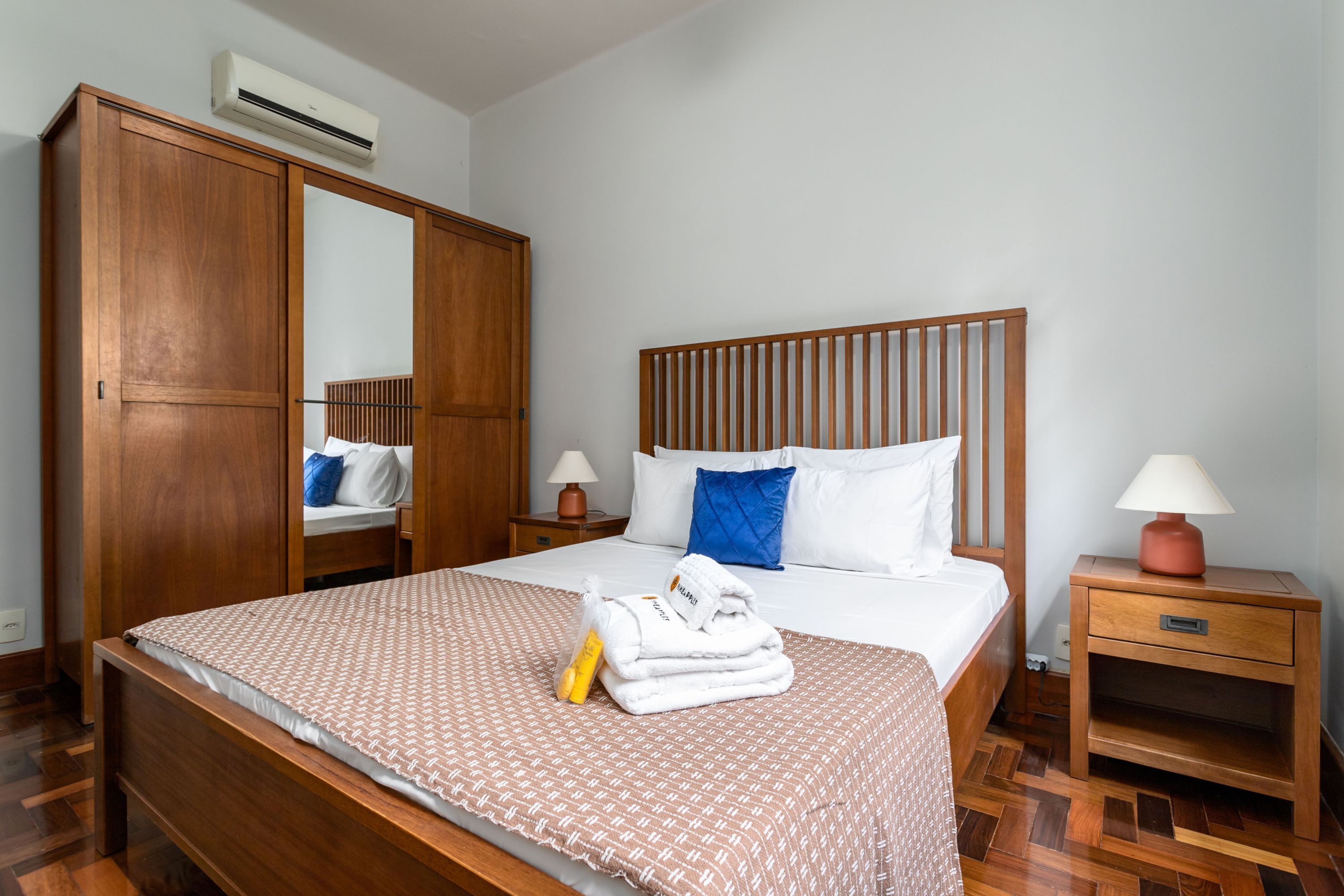 Quarto de hotel com cama de madeira, guarda-roupa, mesa de cabeceira e uma luminária.