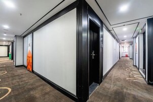 Corredor de hotel com piso carpetado, porta com moldura preta e parede branca com um pôster.