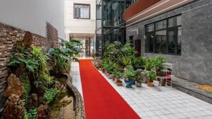 Corredor com carpete vermelho, plantas em vasos e parede de tijolos em um lado.
