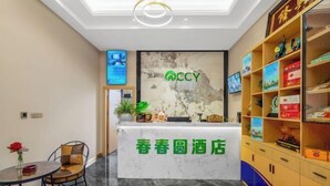Um balcão de recepção com tampos de mármore, um logotipo verde e uma placa com caracteres chineses. À esquerda, há uma pequena mesa com cadeiras e um vaso de flores.