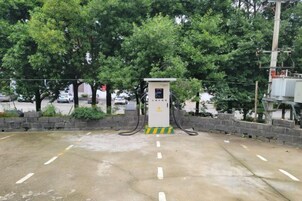 Posto de combustível com uma pequena loja, rodeado por árvores e uma área pavimentada demarcada por linhas brancas.