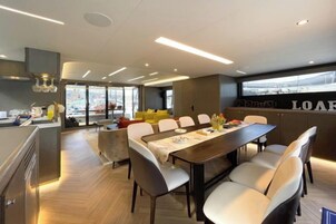Uma área de refeições moderna com uma mesa comprida e cadeiras, cozinha ao fundo e vista através de grandes janelas.
