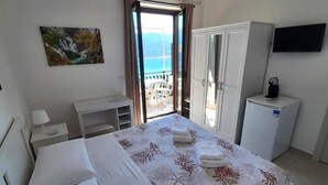 Quarto com cama, escrivaninha, guarda-roupa, televisão e varanda com vista para o mar.