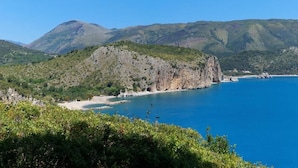 Paisagem costeira com um imponente falcão rochoso, uma praia e mar azul cristalino.