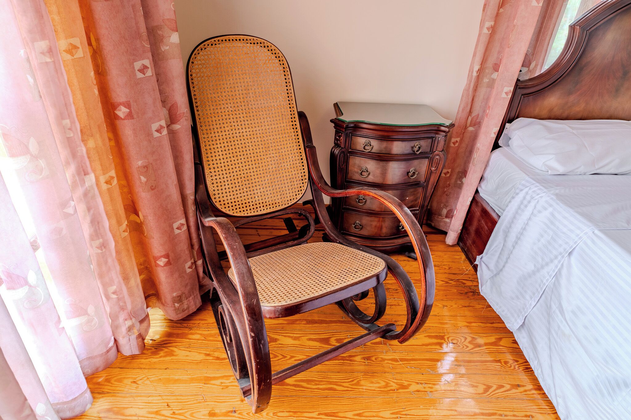 Uma cadeira de balanço de madeira com assento trançado, um criado-mudo de madeira com gavetas e uma cama com roupa de cama branca.