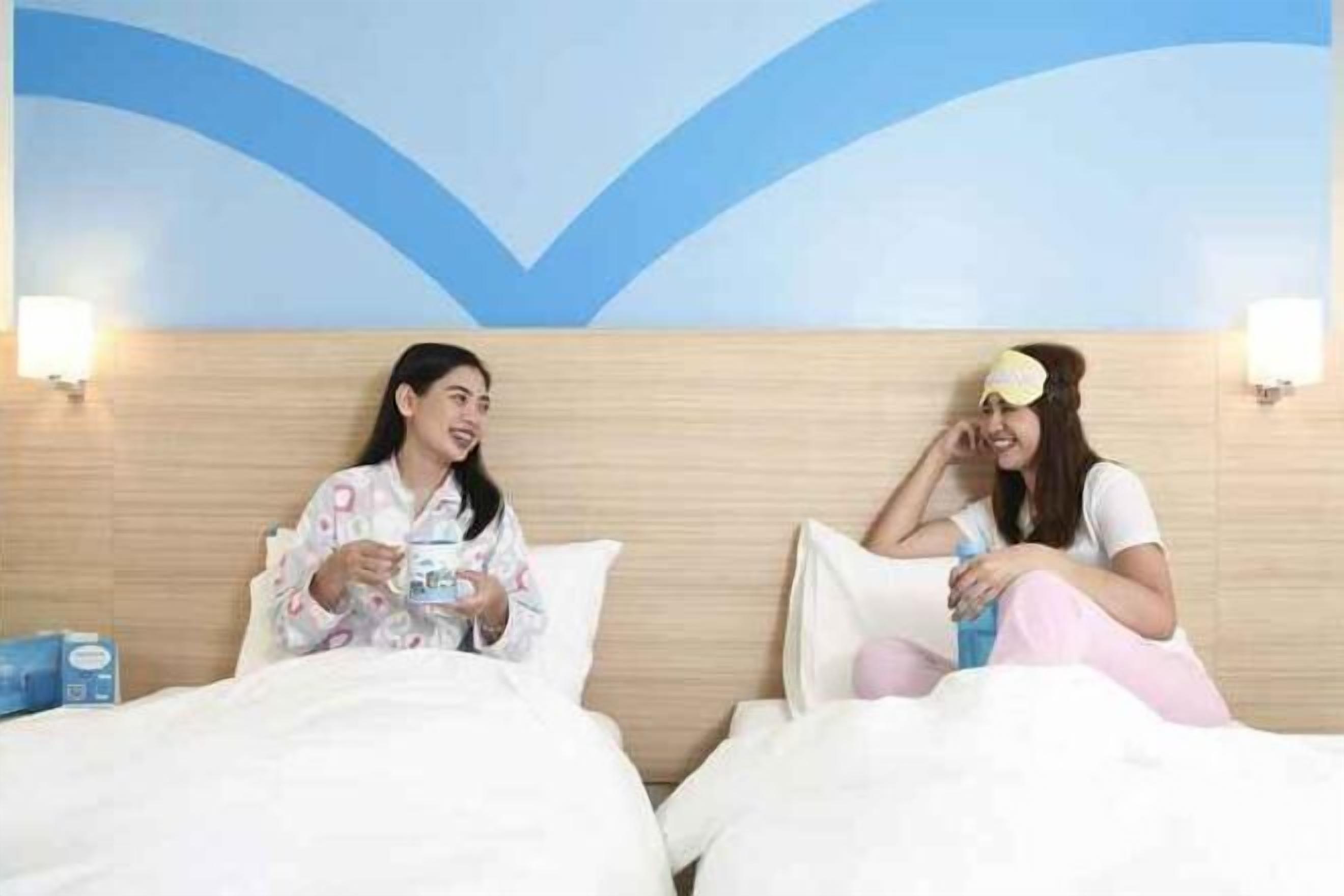 Duas pessoas em quarto de hotel, com cabeceira de madeira, roupa de cama branca e luminárias fixadas na parede.