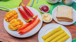 Uma mesa com pratos de manga fatiada, melancia, mamão e enrolados amarelos, juntamente com manteiga e geleia.