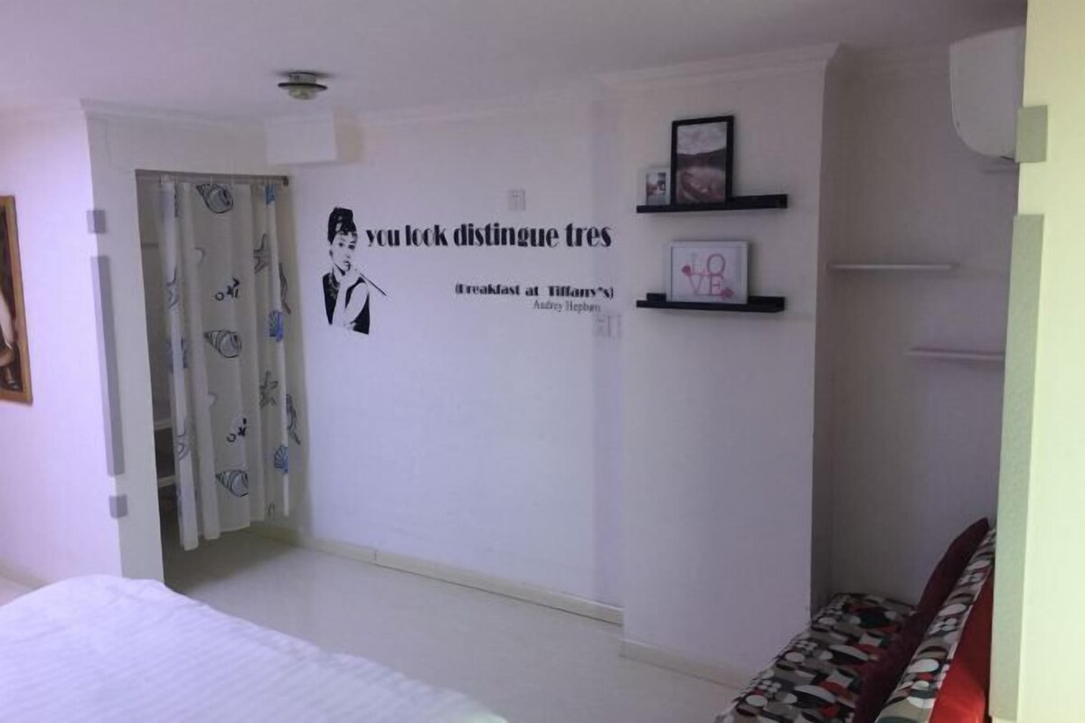 Um quarto com cama, parede com frase inspiradora, porta com cortinas e prateleira com fotografias.
