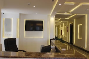 Uma área de recepção de hotel com balcão de mármore, uma cadeira de escritório preta, uma TV de tela plana na parede e um corredor com paredes e portas iluminadas.