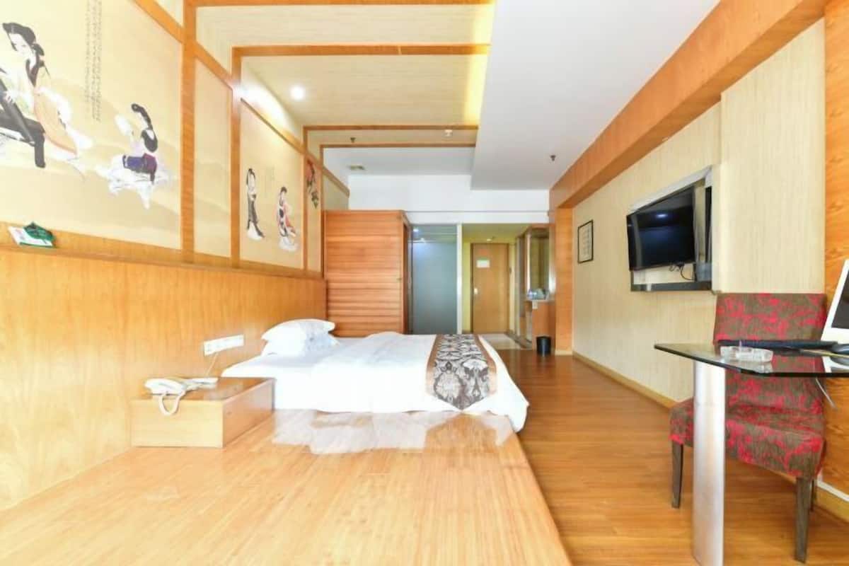 Quarto de hotel com piso de madeira, cama com roupa de cama branca, escrivaninha com computador, televisão e obras de arte nas paredes.