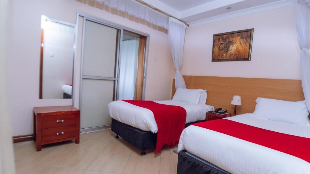 Quarto de hotel com duas camas, uma mesinha de cabeceira de madeira, um espelho, uma porta de correr e um quadro na parede.
