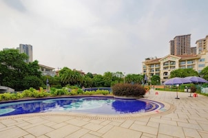 Piscina com deck revestido, rodeada por vegetação e edifícios.