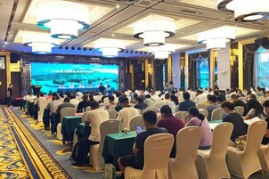 Uma sala de conferências com fileiras de mesas e cadeiras, uma tela grande na frente e um tapete estampado.