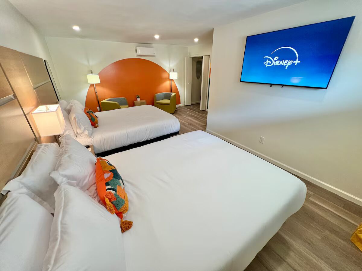 Quarto de hotel com duas camas, uma TV exibindo o logotipo do Disney+ e uma parede curva laranja.